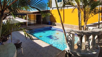 Piscina com deck ladrilhado, rodeada por palmeiras e um prédio com paredes amarelas.