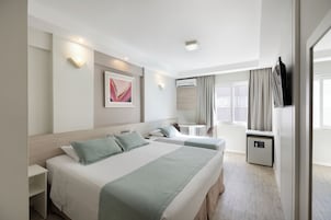 Quarto de hotel com duas camas, uma escrivaninha, uma cadeira, uma mesinha, um guarda-roupa e uma janela com cortinas.
