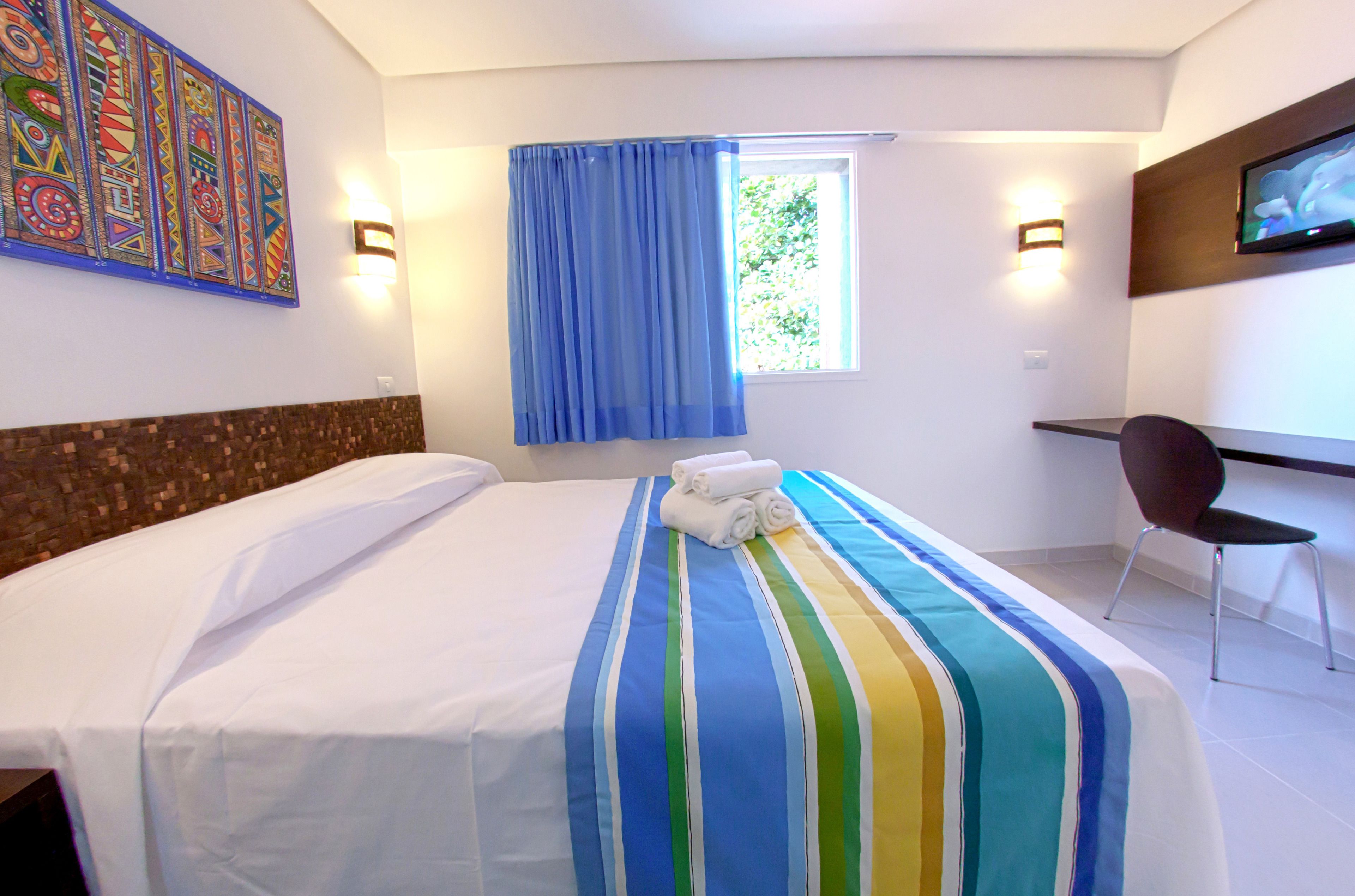 Quarto de hotel com cama, janela com cortinas, televisão fixada na parede e uma cadeira.