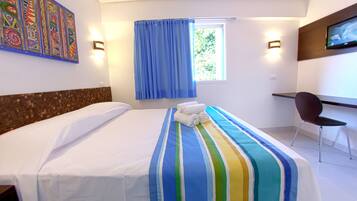 Quarto de hotel com cama, janela com cortinas, televisão fixada na parede e uma cadeira.