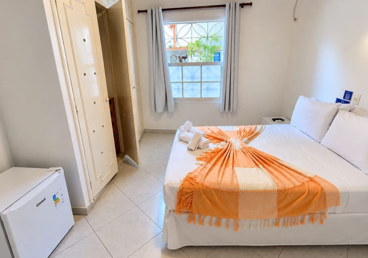Quarto de hotel com cama, mini-frigorífico, janela com cortinas e vista para o verde lá fora.