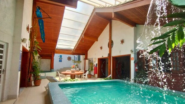 Piscina com cachoeira, rodeada por um alpendre coberto com assentos e plantas em vasos.