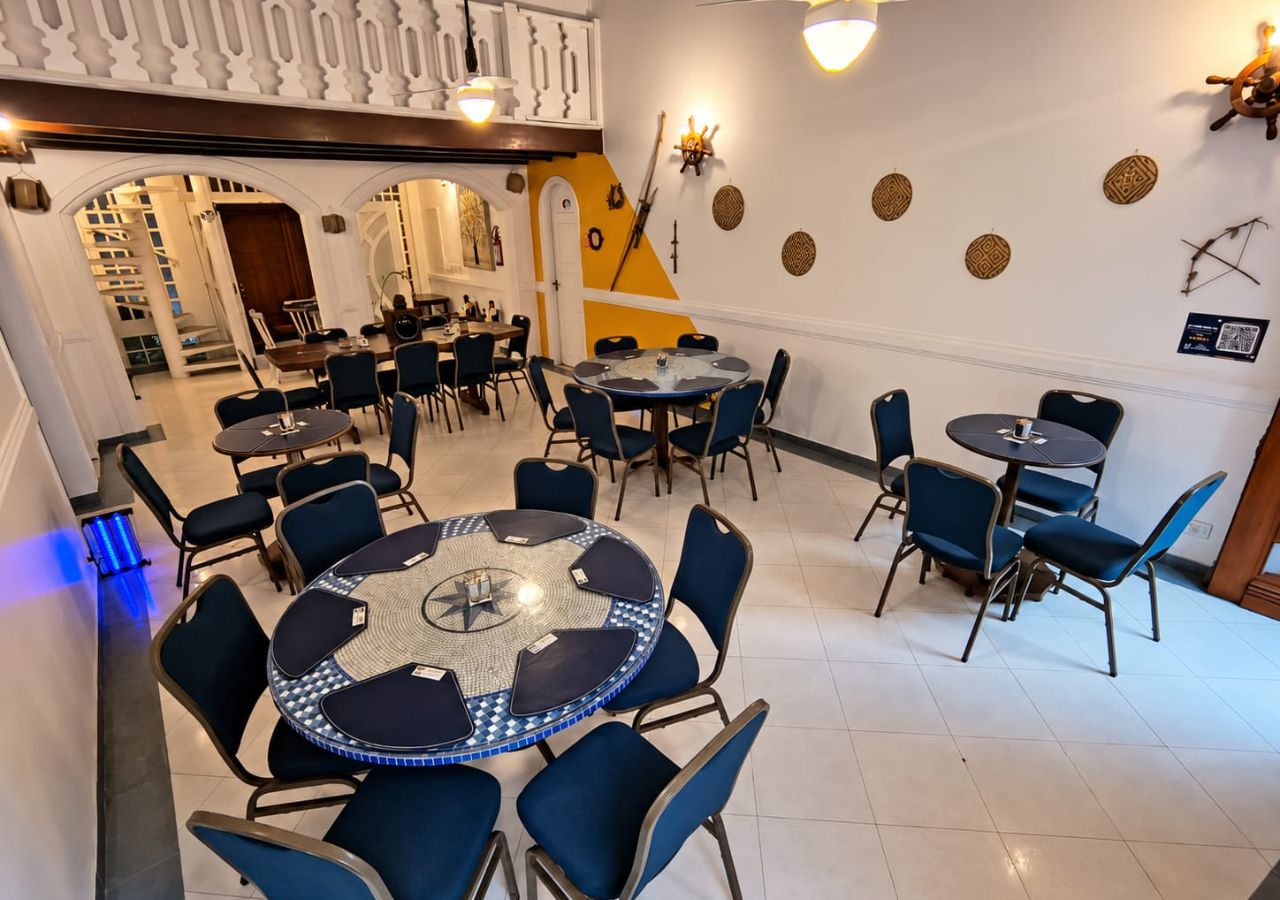 Uma área de refeições de hotel com várias mesas redondas e cadeiras, uma escadaria e detalhes decorativos nas paredes.