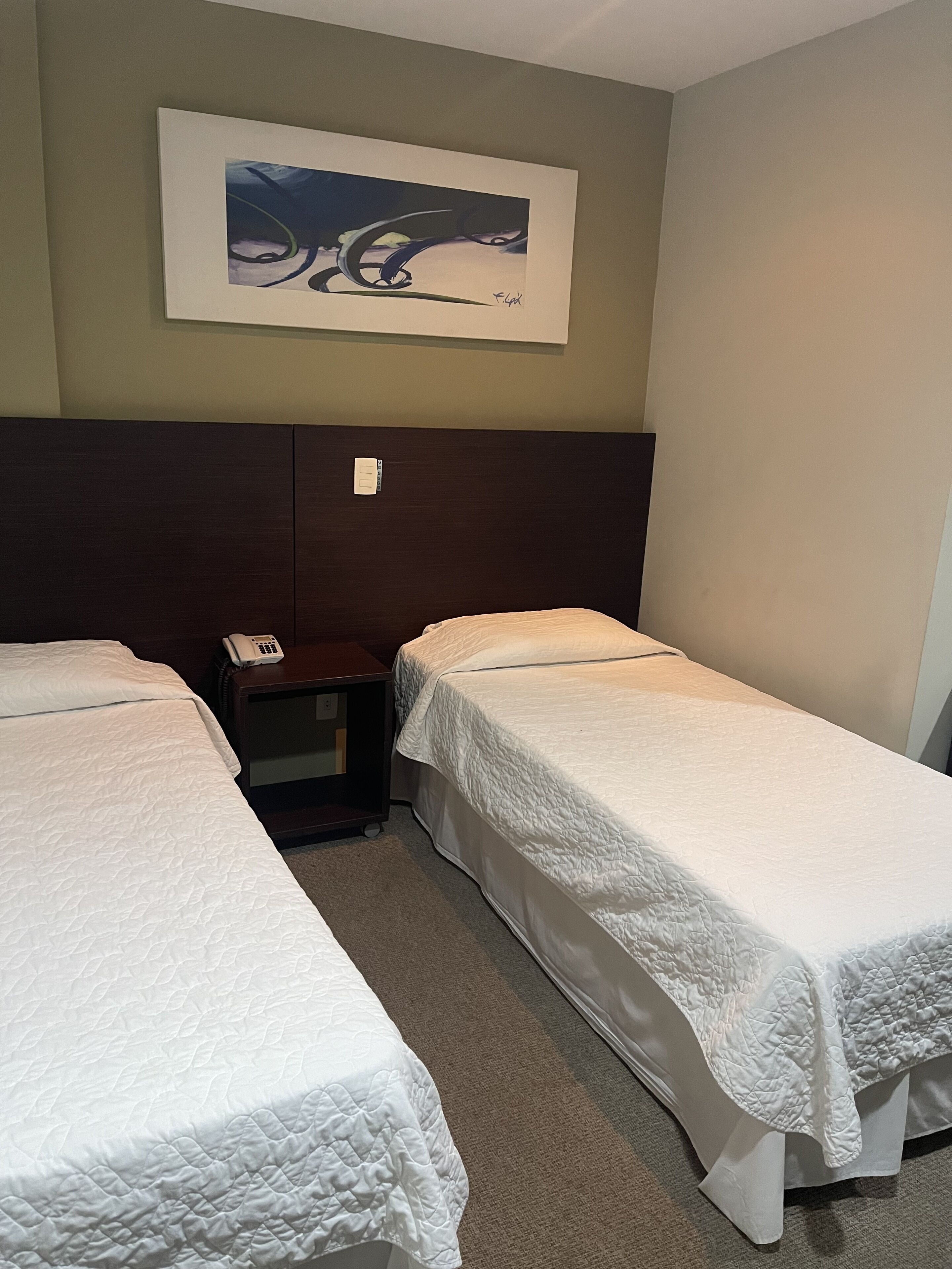 Quarto de hotel com duas camas de solteiro, uma mesinha de cabeceira com telefone e um quadro na parede.