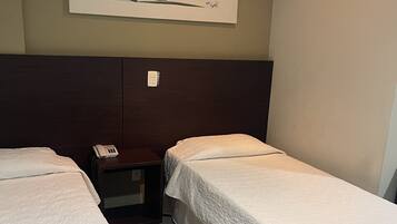 Quarto de hotel com duas camas de solteiro, uma mesinha de cabeceira com telefone e um quadro na parede.
