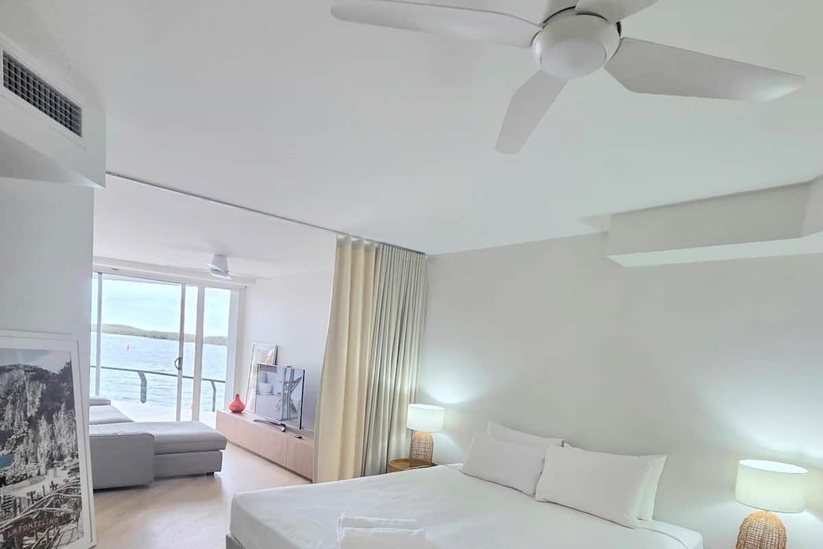 Quarto de hotel moderno com uma cama grande, ventilador de teto, janela com vista para o mar e uma pequena mesa de cabeceira com abajur.