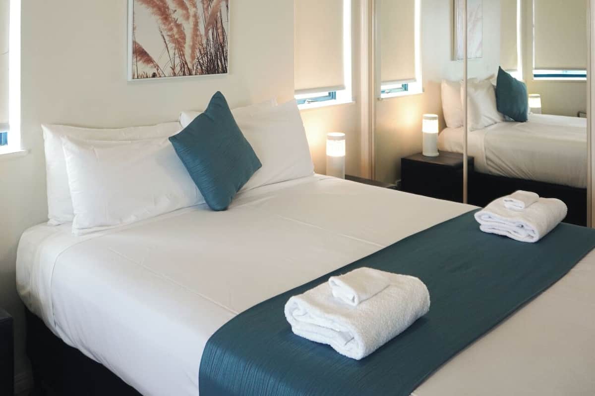 Quarto de hotel com cama, mesa de cabeceira com abajur, espelho e obra de arte na parede.
