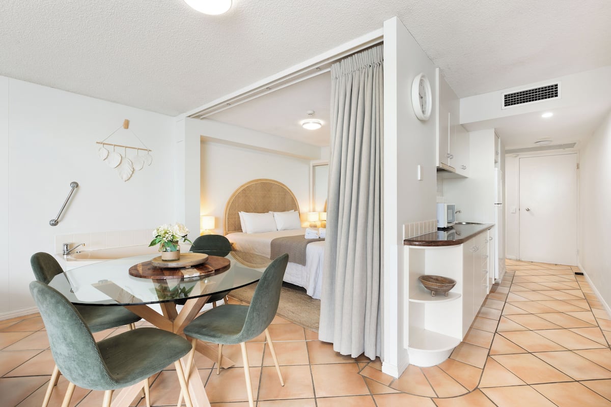Quarto de hotel moderno com mesa de jantar de vidro, cadeiras estofadas verdes, cama com roupa de cama branca, pequena área de cozinha e banheiro com chuveiro.