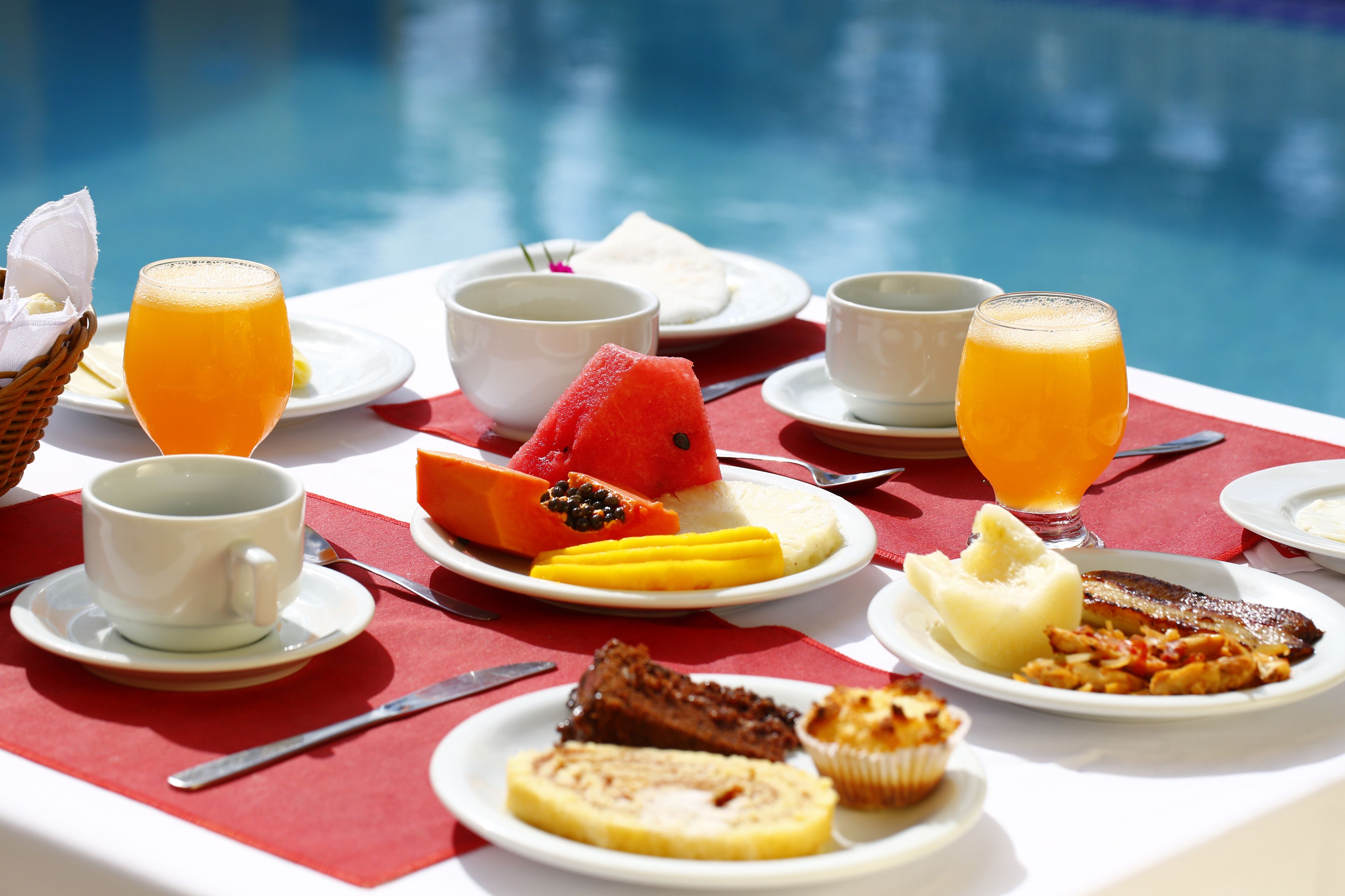 Mesa de café da manhã ao lado de uma piscina, com pratos de comida, xícaras de café e copos de suco de laranja.