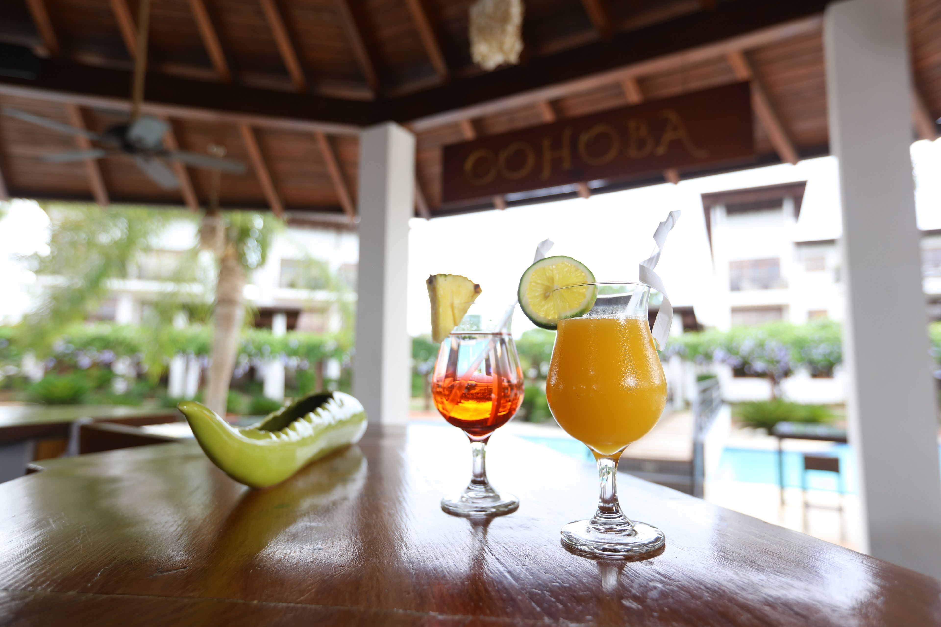 Uma mesa de madeira com três bebidas, uma colher verde e uma placa com a inscrição "COHOBA".