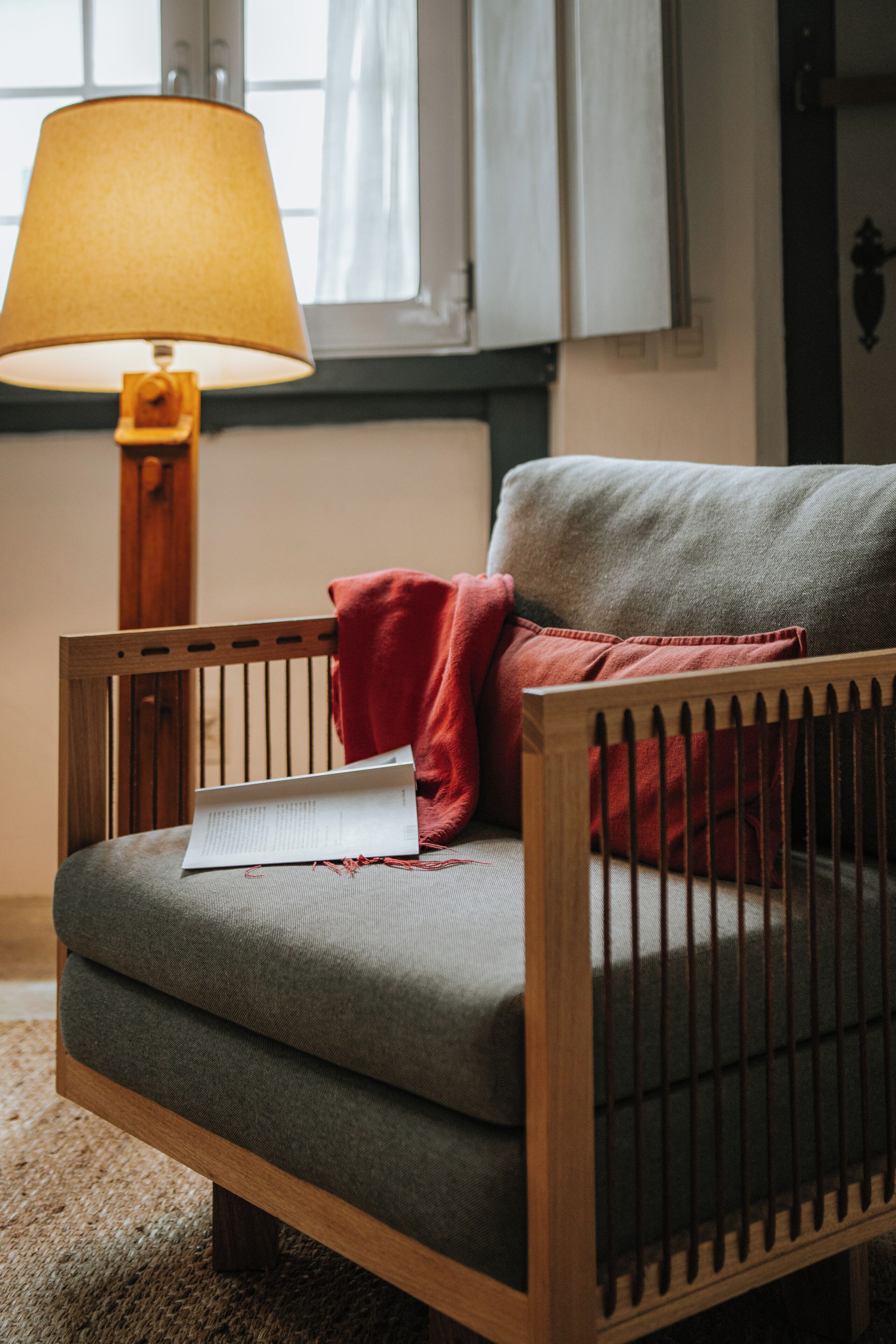 Um berço de madeira com um travesseiro cinza e um vermelho, uma luminária amarela e uma janela com cortinas ao fundo.