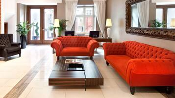 Uma sala de estar moderna com um sofá vermelho, uma poltrona preta e uma mesa de centro de madeira. Há duas luminárias em uma mesinha lateral e um espelho grande na parede.