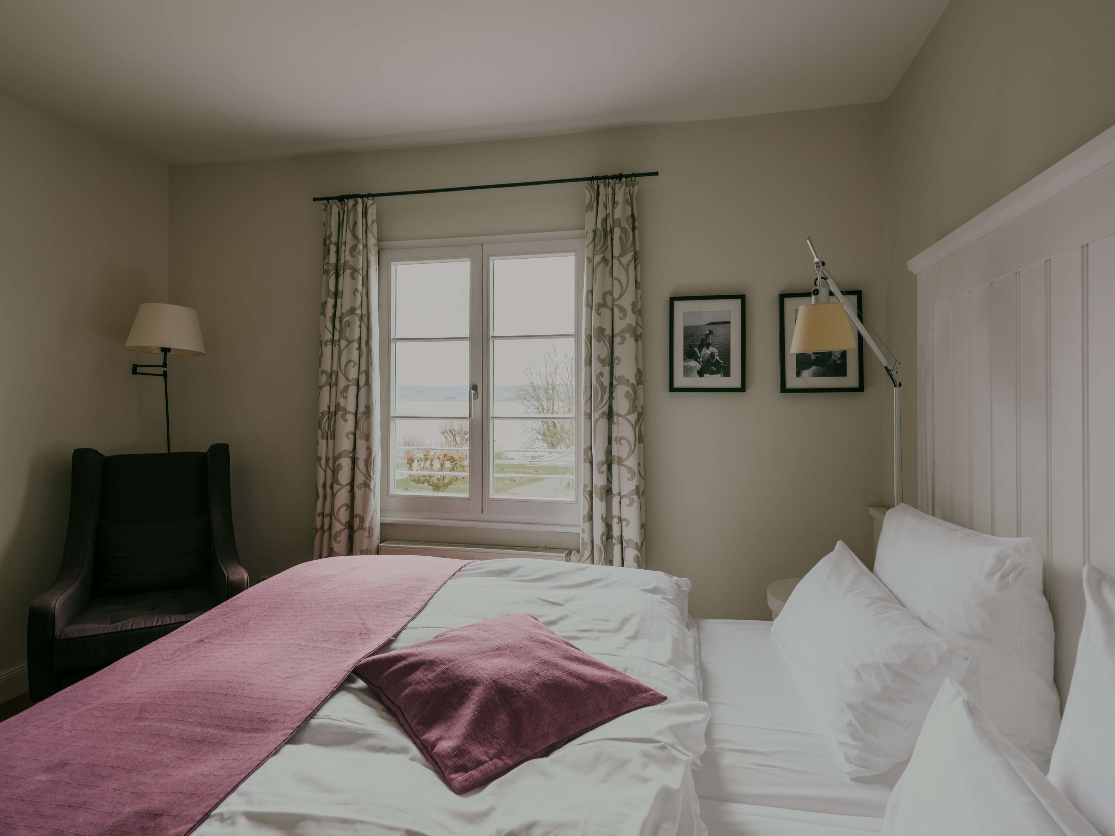 Quarto com cama, uma cadeira, janela com cortinas e duas luminárias na parede.