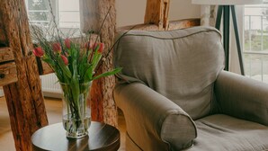 Quarto com vigas de madeira aparentes, um sofá cinza, uma mesa redonda de madeira com um vaso de flores e um abajur.