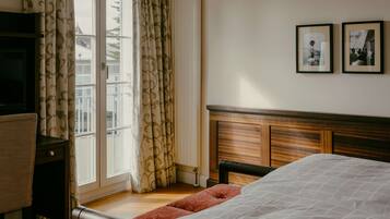 Quarto de hotel com cama, escrivaninha, cadeira, janela com cortinas e duas imagens emolduradas na parede.