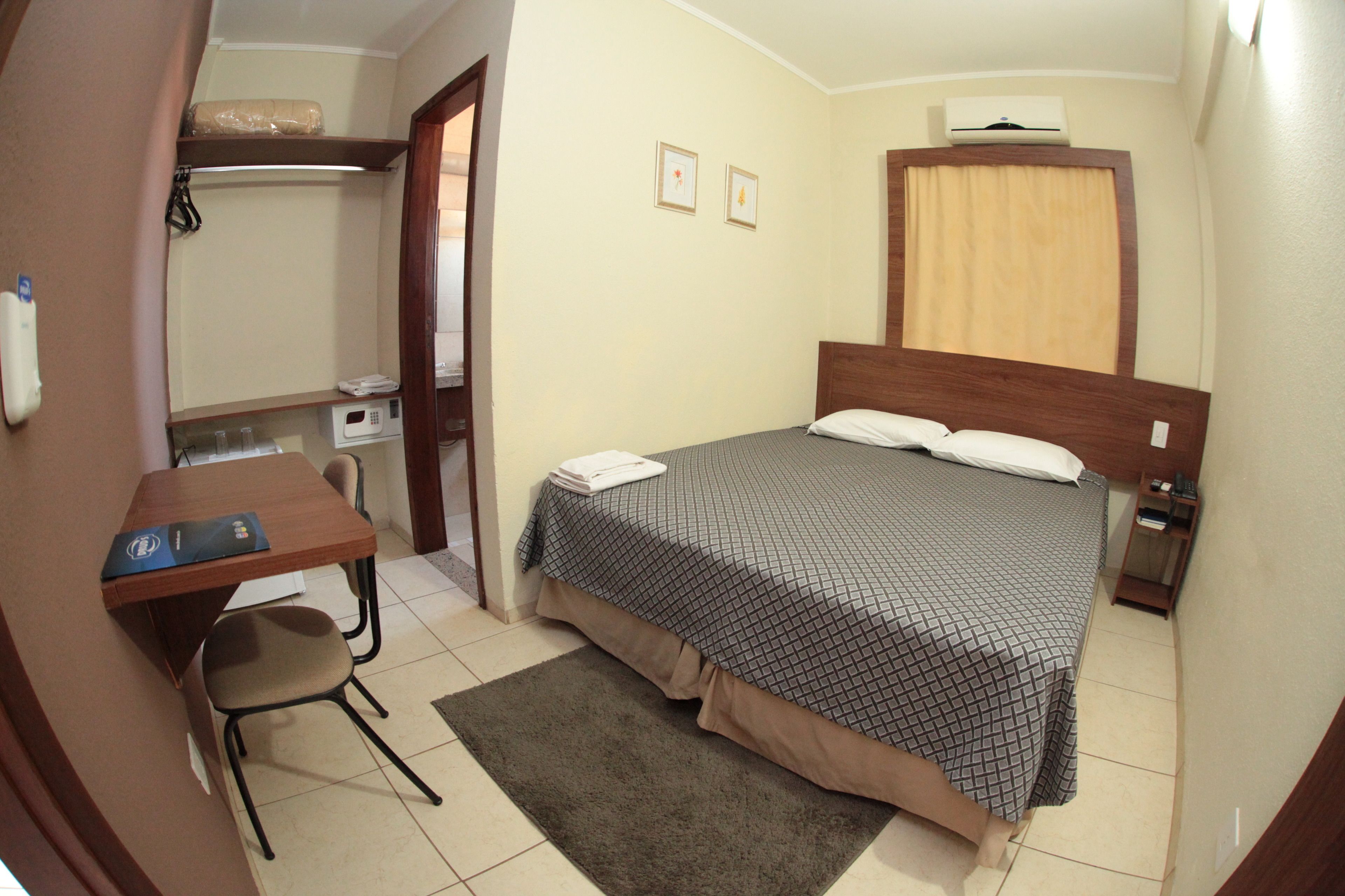 Quarto de hotel com cama, escrivaninha com cadeira, mesa pequena, micro-ondas e armário.
