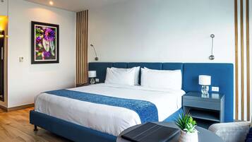 Quarto de hotel moderno com uma cama grande, mesa de cabeceira, luminária, uma pequena planta e um laptop sobre uma mesa redonda.