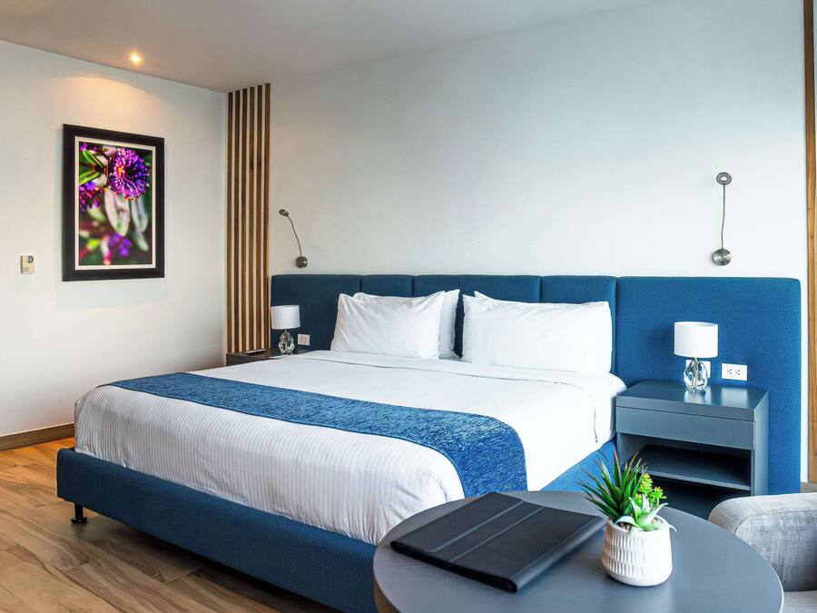 Quarto de hotel moderno com uma cama grande, mesa de cabeceira, luminária, uma pequena planta e um laptop sobre uma mesa redonda.