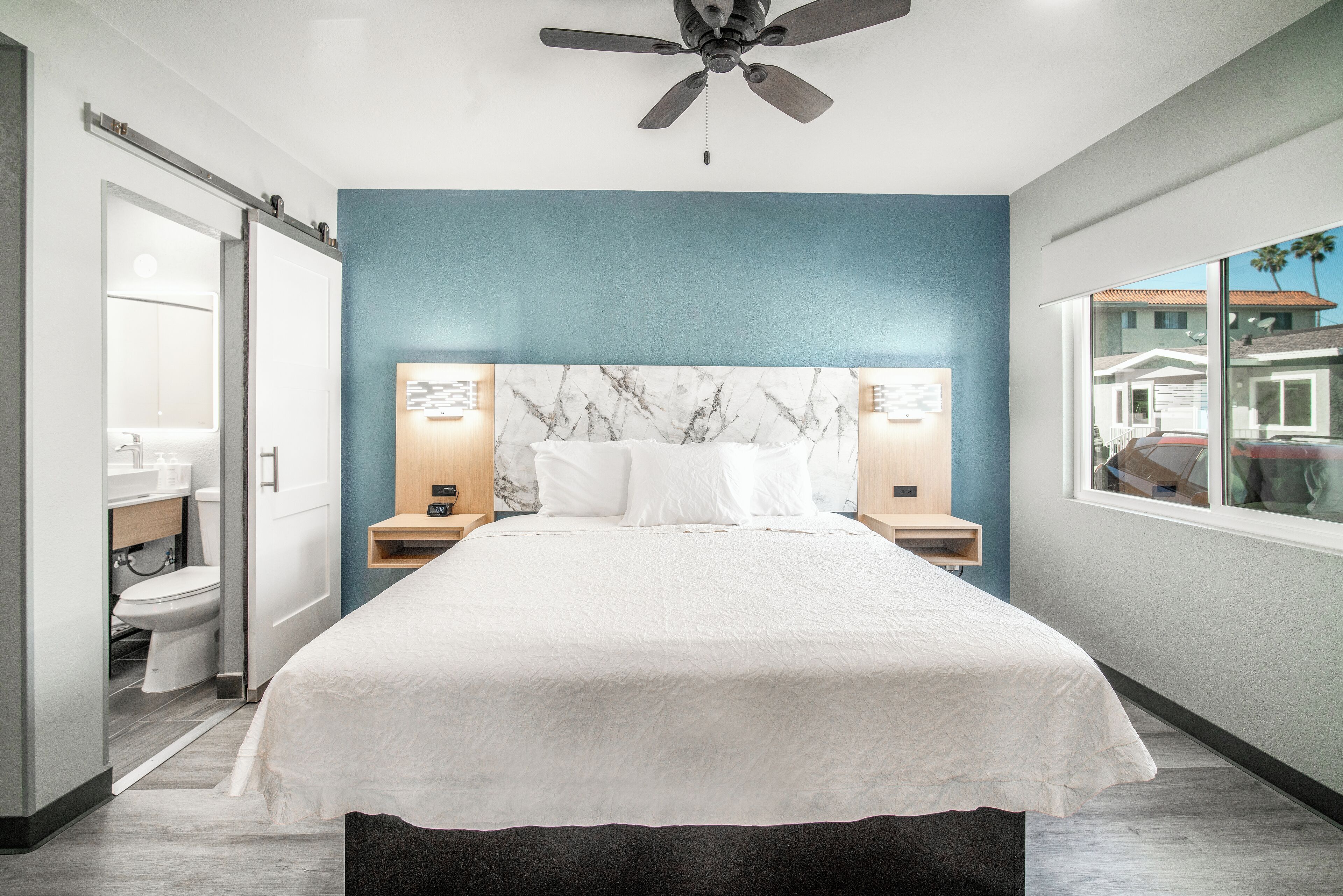 Quarto com cama, ventilador de teto, cabeceira de mármore, abajures e janela com vista para prédios.