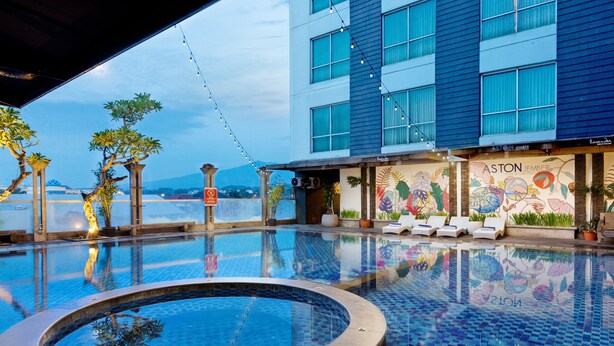 Área de piscina de hotel com vista para uma montanha, cadeiras de descanso e um mural no prédio.