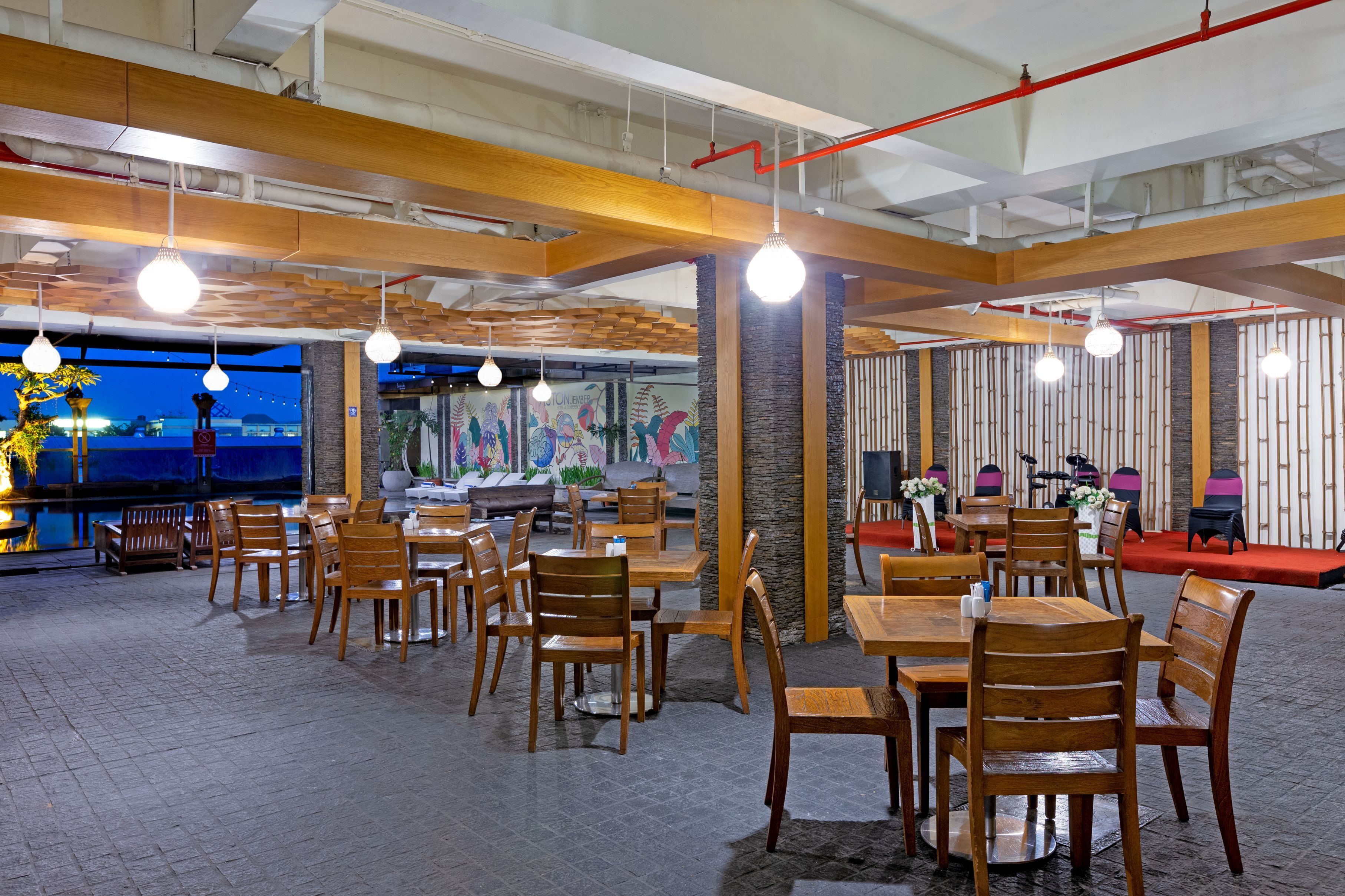 Um restaurante amplo com mesas e cadeiras de madeira, luminárias pendentes e vista para a cidade através de grandes janelas.