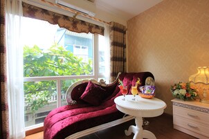 Quarto com uma grande janela, um sofá vermelho confortável, uma mesinha redonda com bebidas e um abajur decorativo.