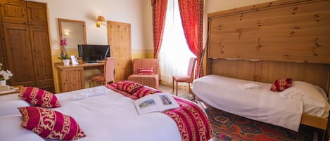 Quarto de hotel com duas camas, um guarda-roupa de madeira, uma mesa de trabalho com televisão e uma janela grande com cortinas vermelhas.