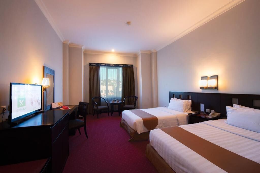 Quarto de hotel com duas camas, uma escrivaninha com computador, uma cadeira e uma janela com cortinas.