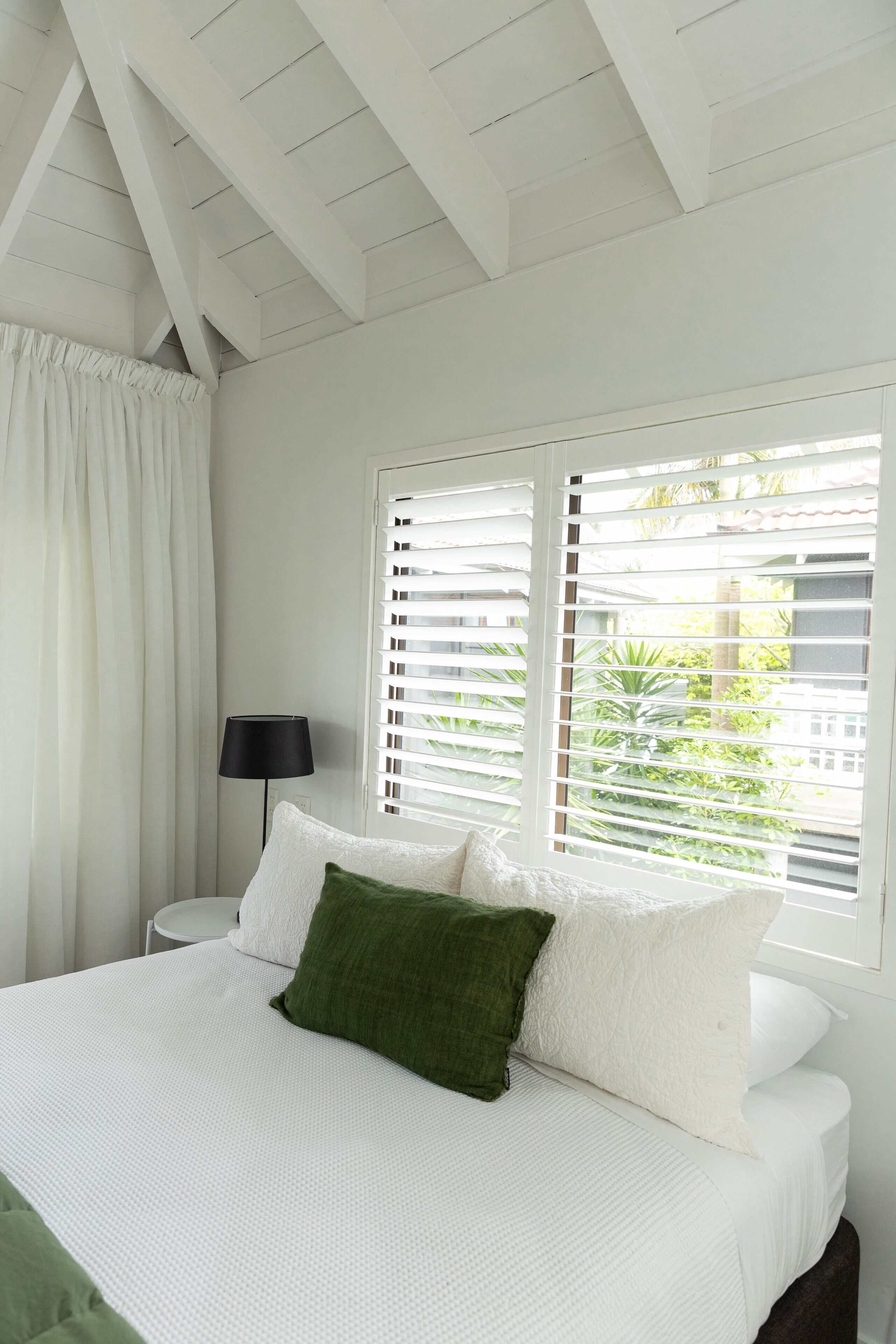 Quarto com cama, cortinas brancas, janela com persianas e mesa de cabeceira com abajur.