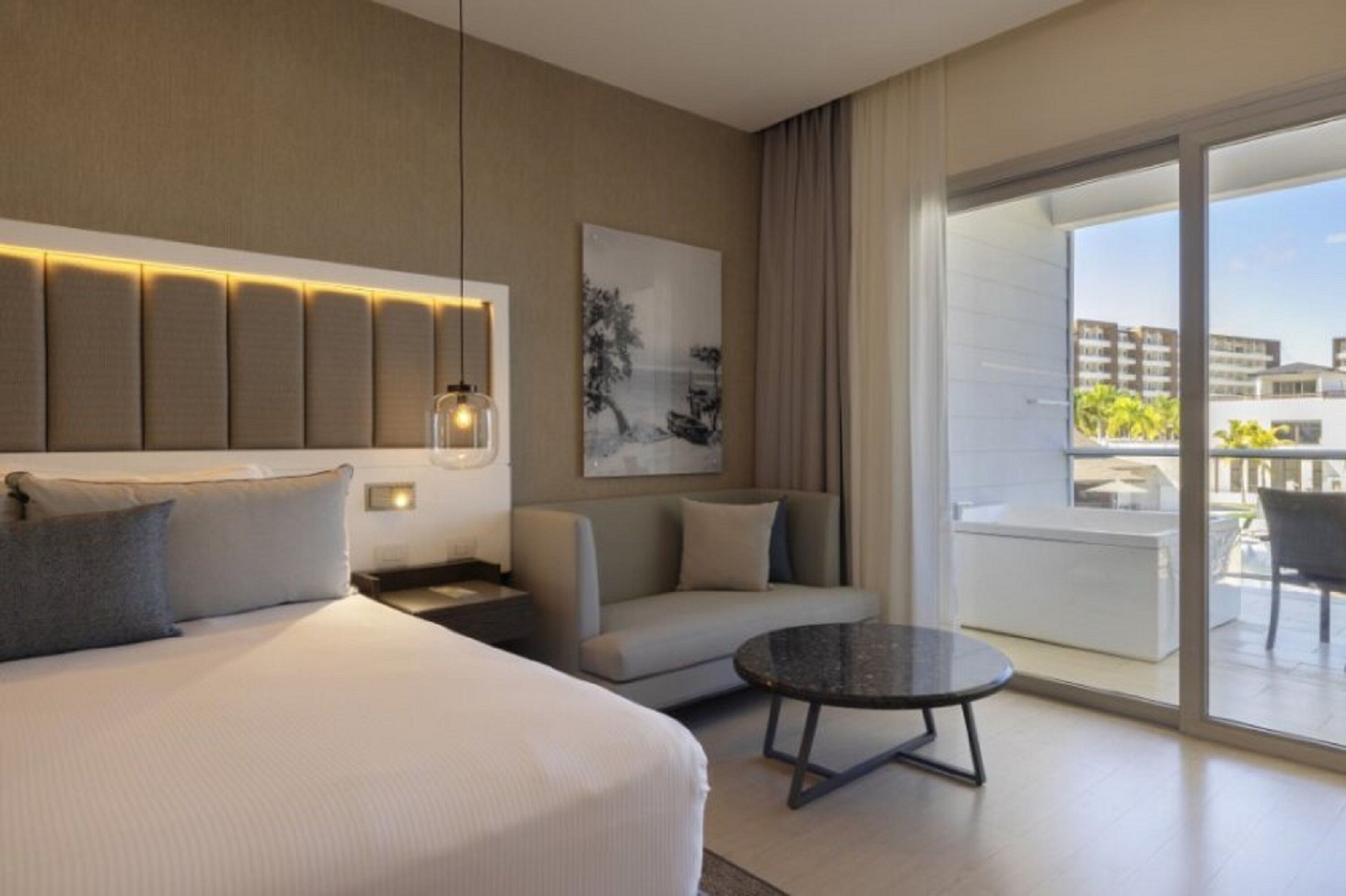 Quarto de hotel moderno com uma cama grande, um sofá, uma pequena mesa redonda e uma varanda com vista para edifícios e vegetação.