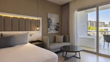 Quarto de hotel moderno com uma cama grande, um sofá, uma pequena mesa redonda e uma varanda com vista para edifícios e vegetação.