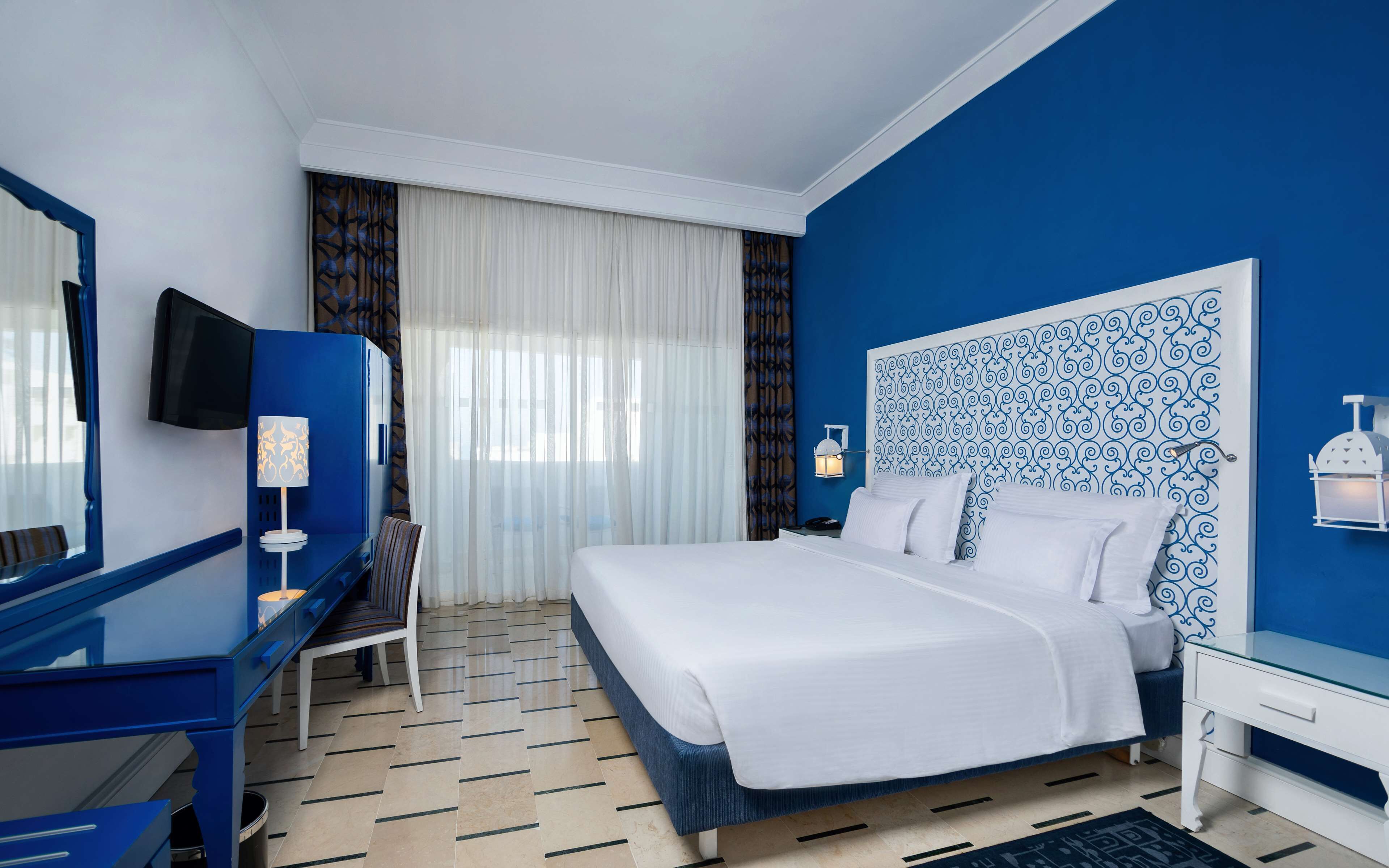 Quarto de hotel com uma cama grande, uma escrivaninha, uma cadeira, uma televisão e uma cabeceira estampada.