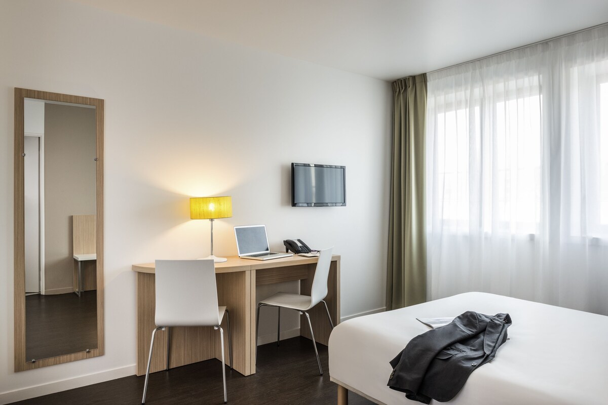 Quarto de hotel com cama, escrivaninha com laptop, cadeira, abajur, telefone, televisão e espelho.