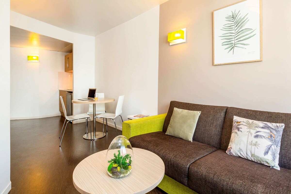 Uma sala de estar moderna com um sofá marrom, uma mesa de centro redonda e uma área de jantar com um laptop e cadeiras.