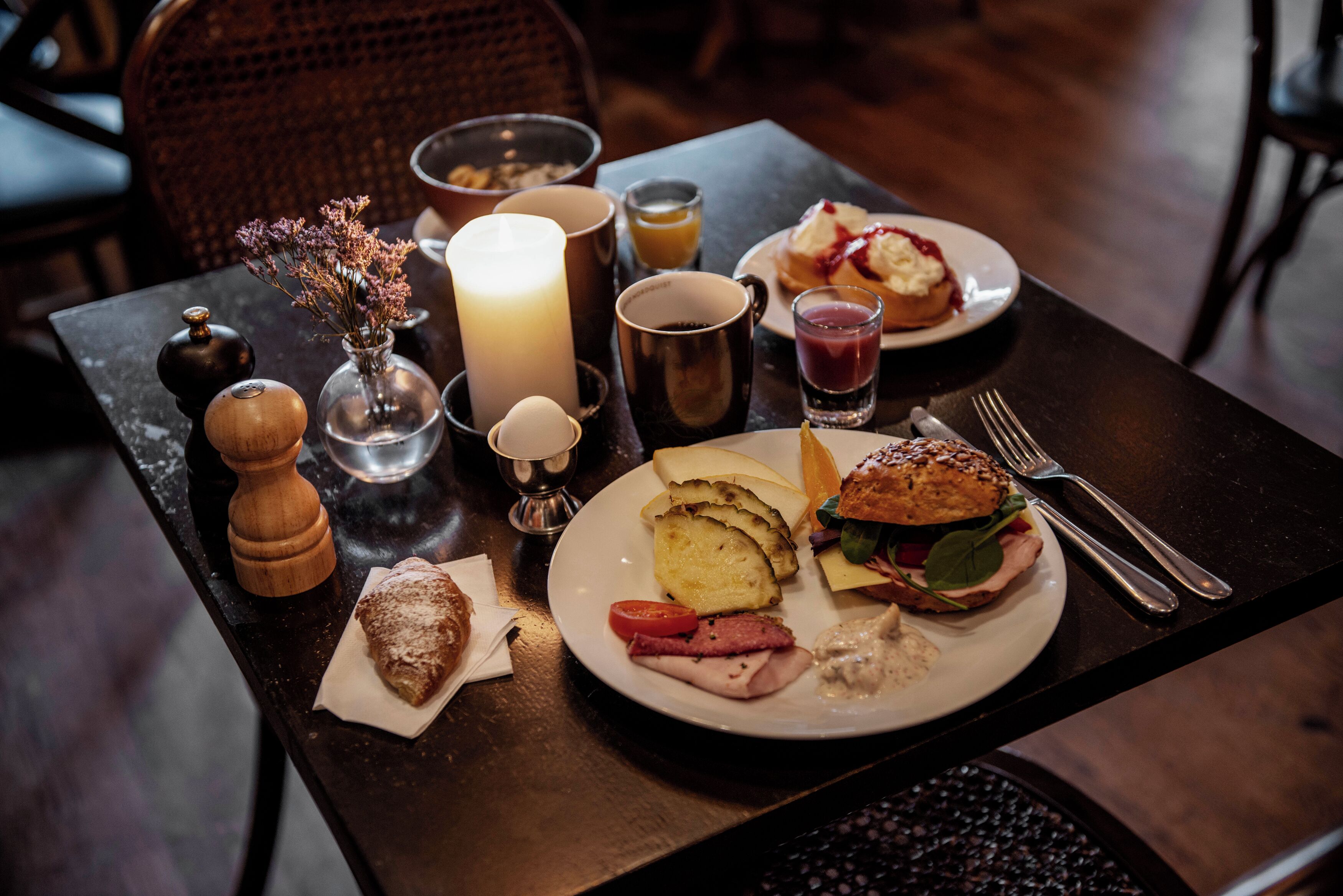 Mesa posta para o café da manhã, com ovos, torradas e um sanduíche, juntamente com uma vela, um saleiro e um pequeno vaso com flores.