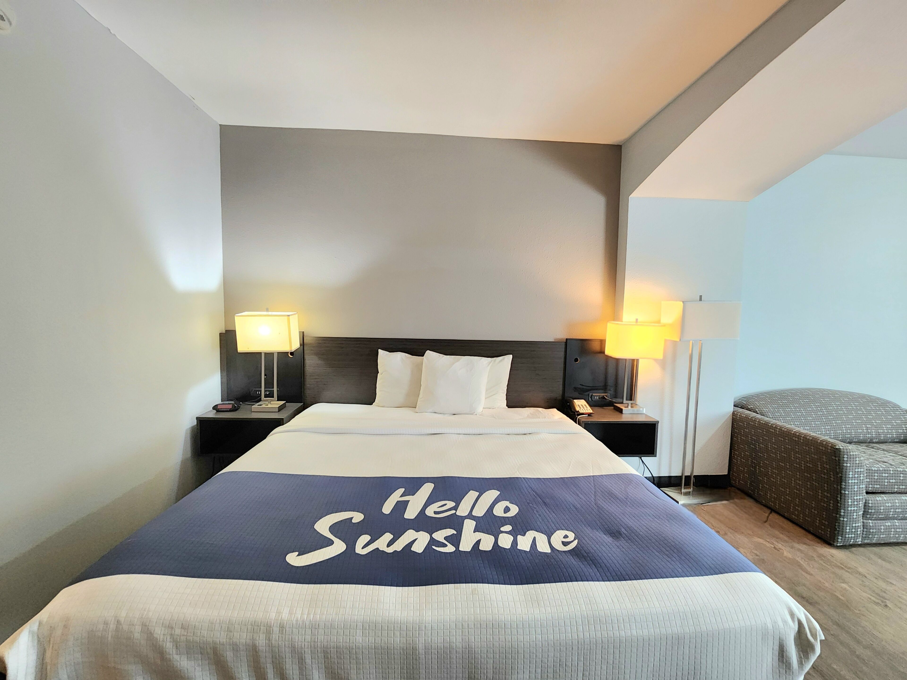 Quarto de hotel com uma cama, duas luminárias laterais, uma cadeira e uma parede com a inscrição 'Hello Sunshine'.