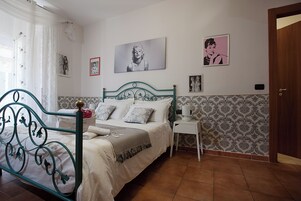 Quarto com cama de estrutura metálica, roupa de cama branca e mesa de cabeceira com abajur. Há quadros nas paredes e uma porta à direita.
