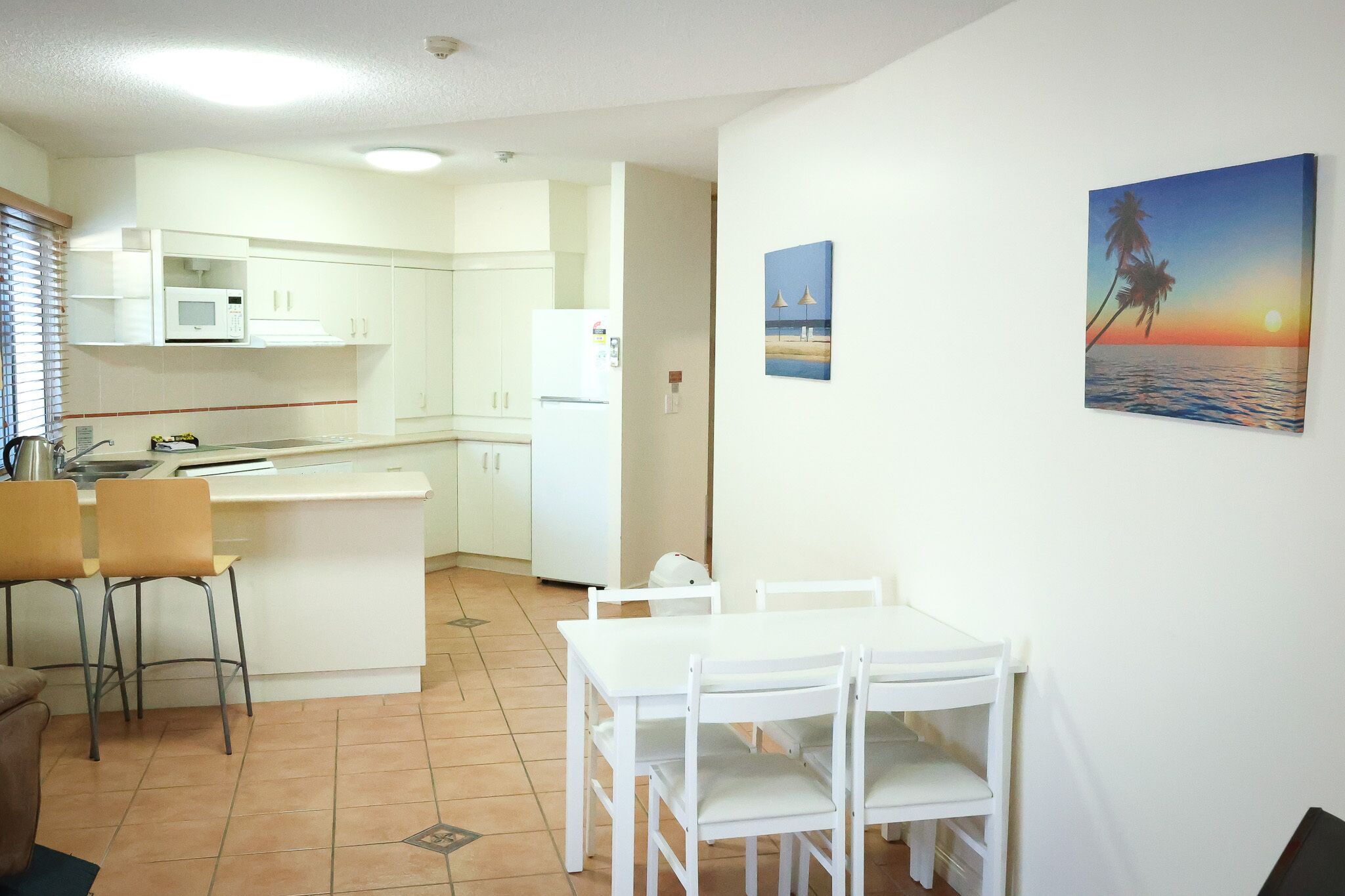 Cozinha compacta com armários brancos, micro-ondas e geladeira. Possui um espaço de jantar com mesa e cadeiras brancas.