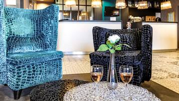 Uma área de recepção com duas cadeiras estampadas, uma mesa redonda pequena com dois copos de vinho e um vaso com uma flor branca.