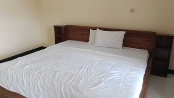 Cama bem arrumada com cabeceira de madeira e roupa de cama branca em um quarto simples, com paredes lisas.