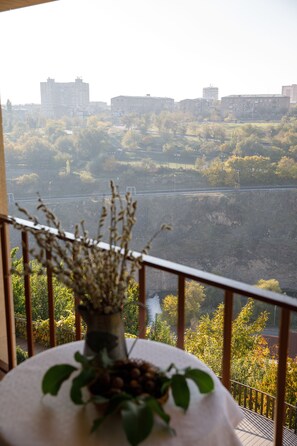 Varanda com mesa onde se encontra um vaso de plantas secas, com vista para um horizonte urbano com edifícios e um rio.