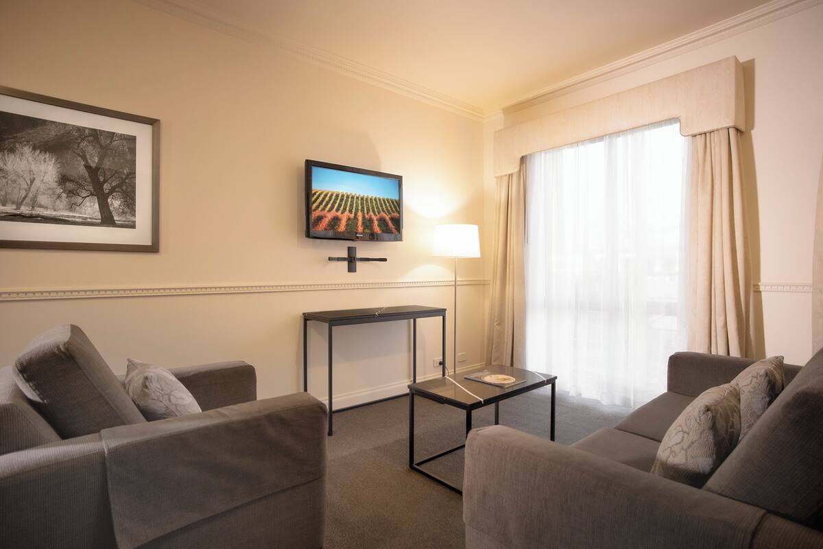 Quarto de hotel com sofá, uma mesinha, televisão, abajur e janela com cortinas.