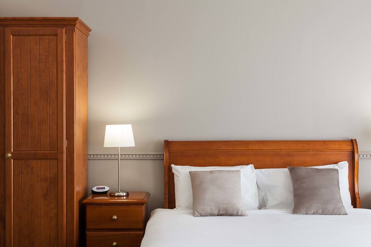 Quarto de hotel com guarda-roupa de madeira, mesa de cabeceira com abajur e cama com duas almofadas.