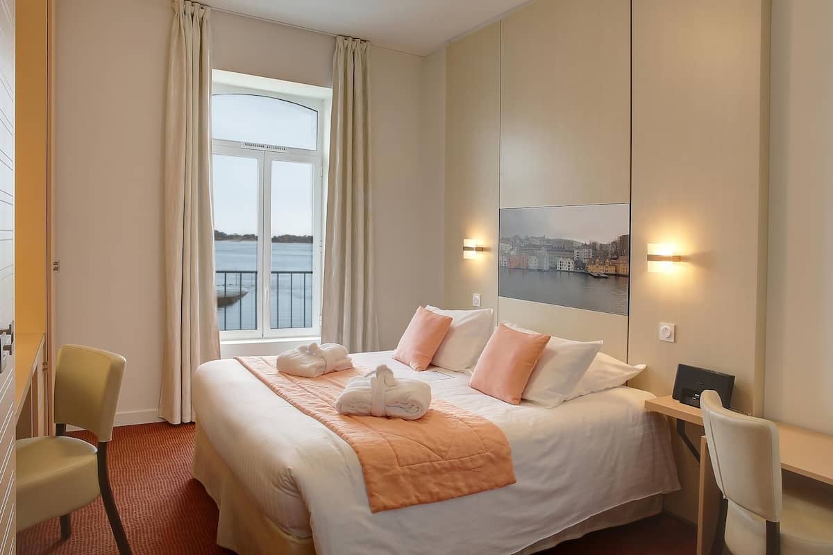 Quarto de hotel com cama, escrivaninha, cadeira, janela com cortinas e um quadro na parede.