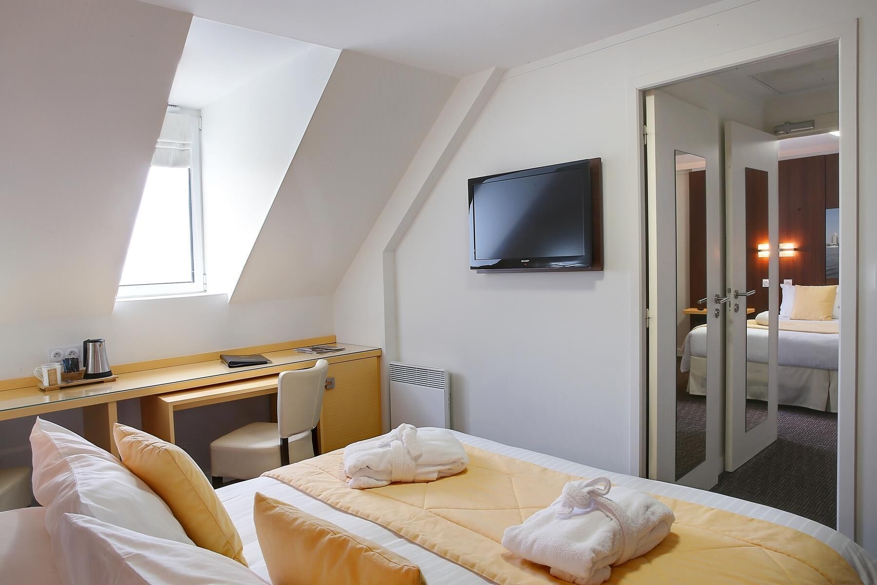 Quarto de hotel com cama, escrivaninha, cadeira, televisão e porta deslizante que dá para outro cômodo.