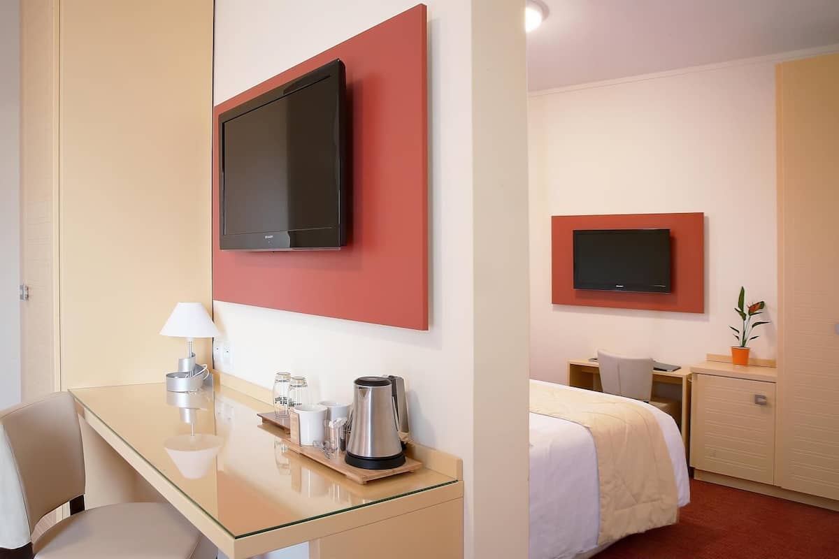 Quarto de hotel com cama, escrivaninha com luminária, cafeteira e duas televisões de tela plana fixadas na parede.