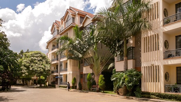 Hotel com vários andares, varandas, palmeiras e um caminho de acesso pavimentado.