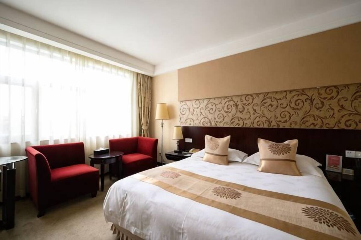 Quarto de hotel com uma cama grande, duas poltronas vermelhas, uma mesa pequena, uma mesinha de cabeceira com abajur e uma cabeceira decorativa.