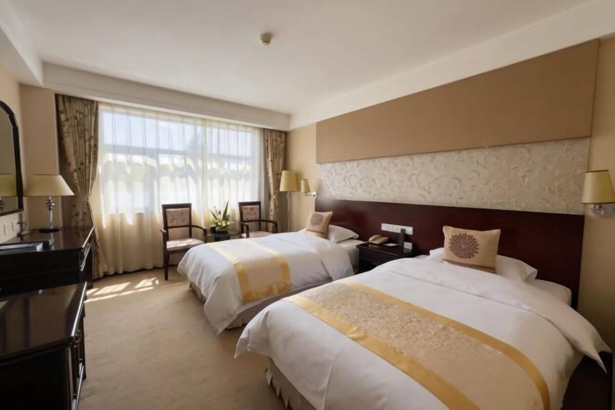 Quarto de hotel com duas camas, uma escrivaninha, uma cadeira, uma mesa de cabeceira e uma janela grande com cortinas.