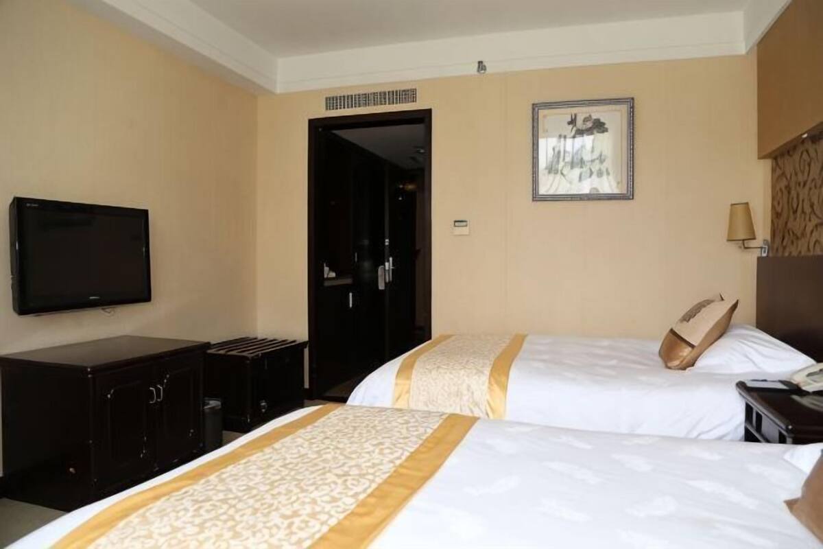 Quarto de hotel com duas camas, uma televisão, uma escrivaninha e uma porta que dá para um corredor.